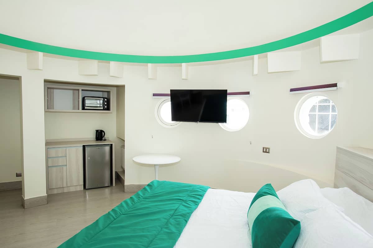 Quarto de hotel moderno com uma cama, um pequeno espaço de cozinha, uma TV de tela plana e duas janelas.
