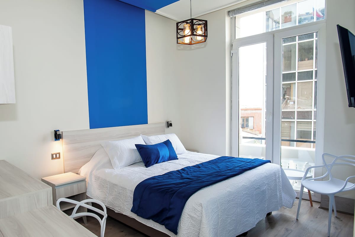 Quarto moderno com uma cama grande, uma mesinha de cabeceira, uma cadeira e vista para o exterior através de uma porta de vidro.