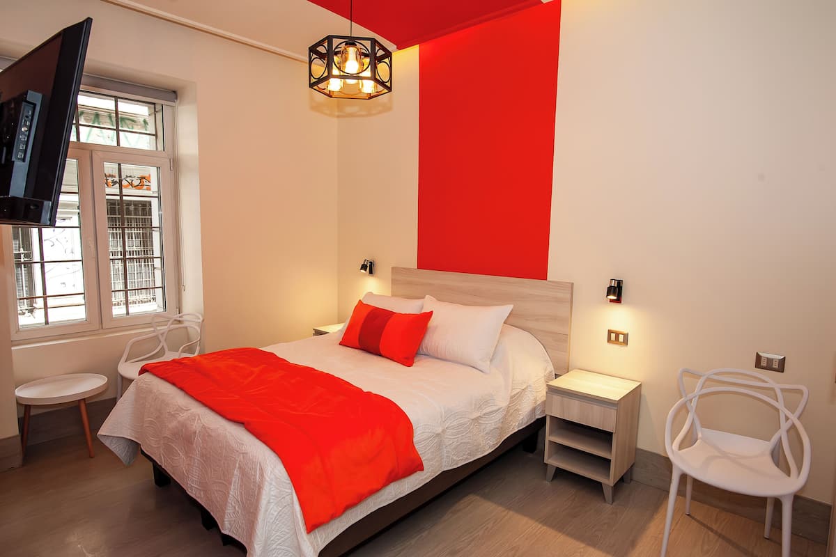Quarto com cama, mesas de cabeceira, cadeira, televisão e janela.