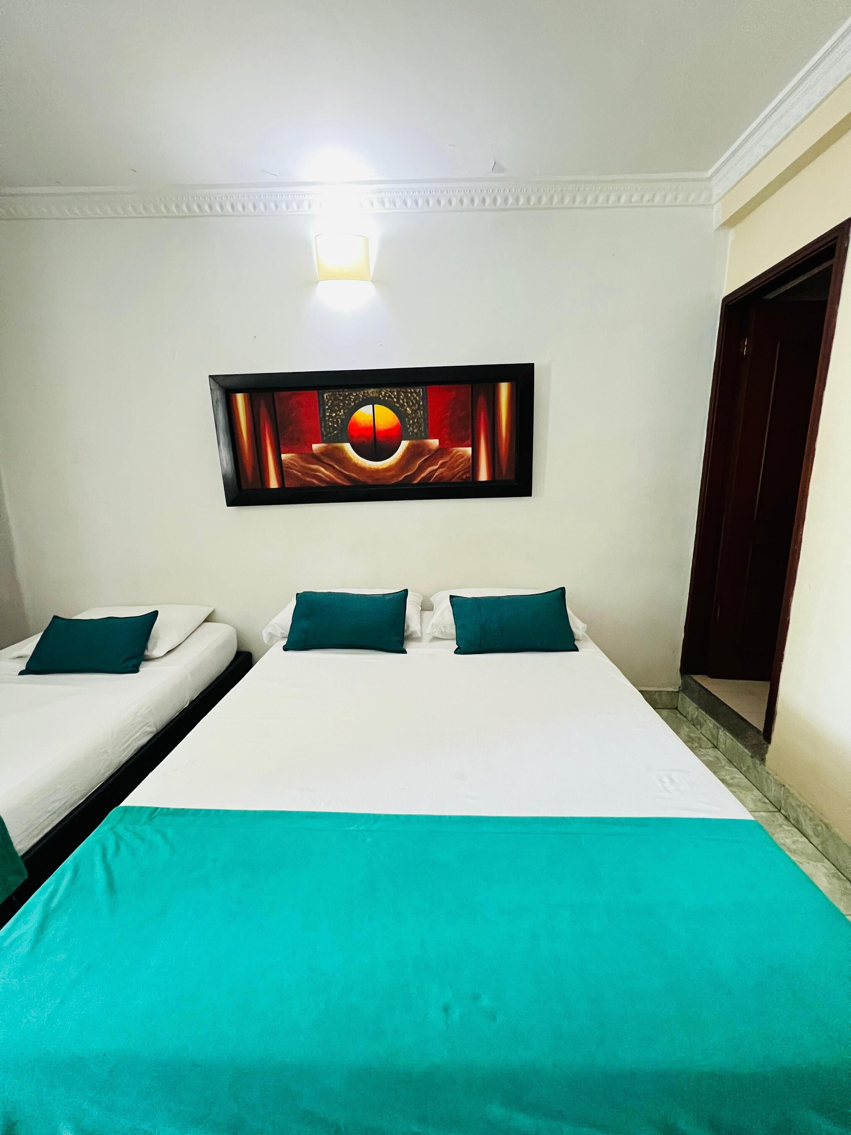 Quarto de hotel com duas camas, roupa de cama azul-turquesa, um quadro sobre as camas e uma porta de madeira.