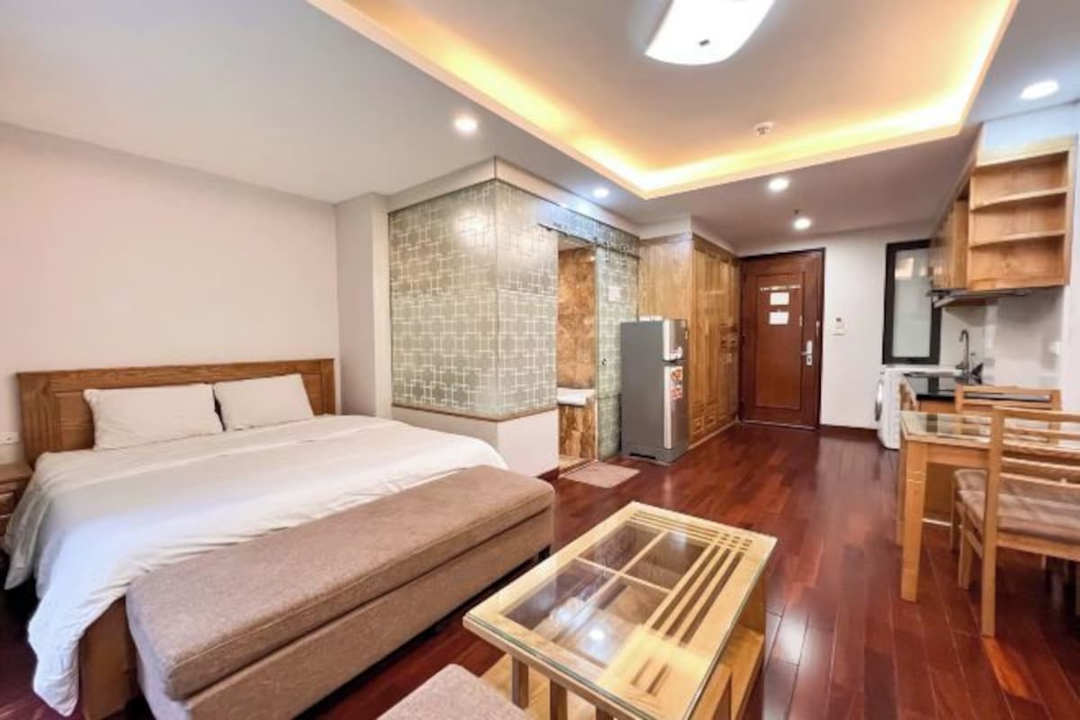 Quarto de hotel com uma cama, mesa com tampo de vidro, cozinha compacta e área de refeições em madeira.