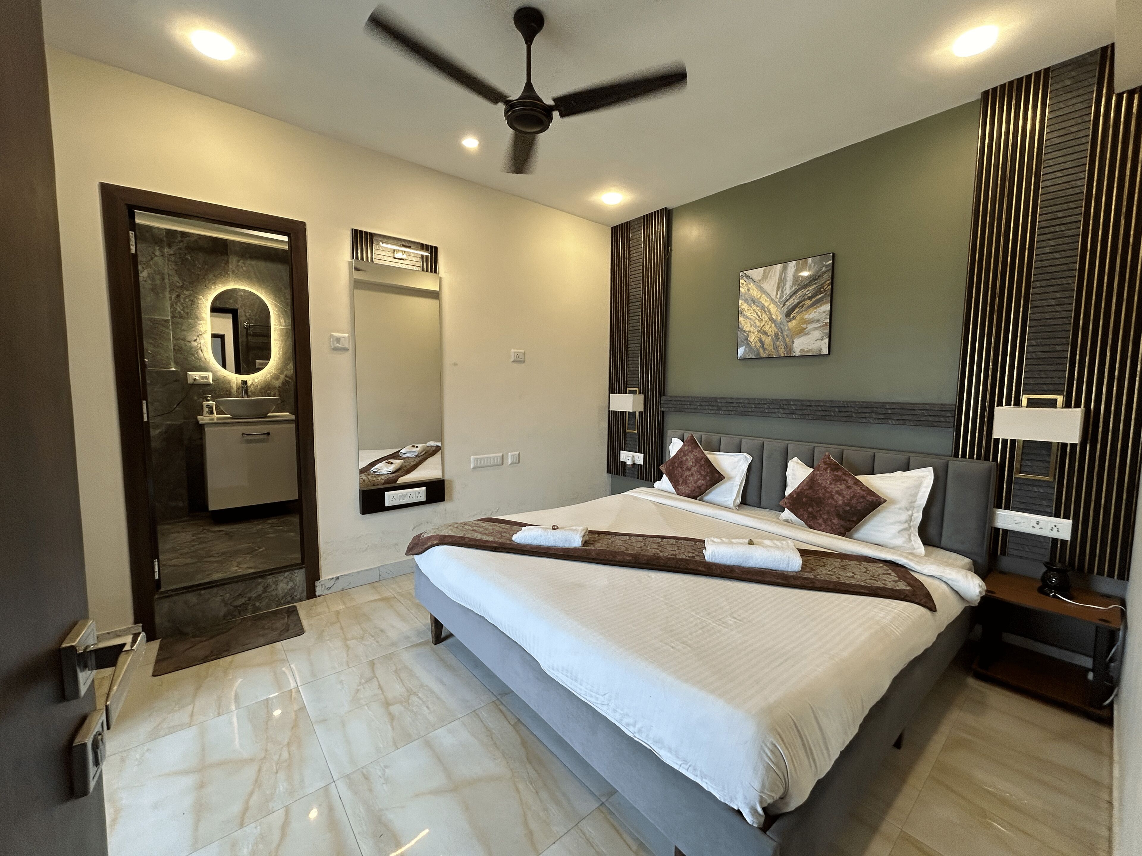 Quarto de hotel moderno com uma cama grande, ventoinha no teto, banheiro com espelho e pia, e um painel decorativo na parede.