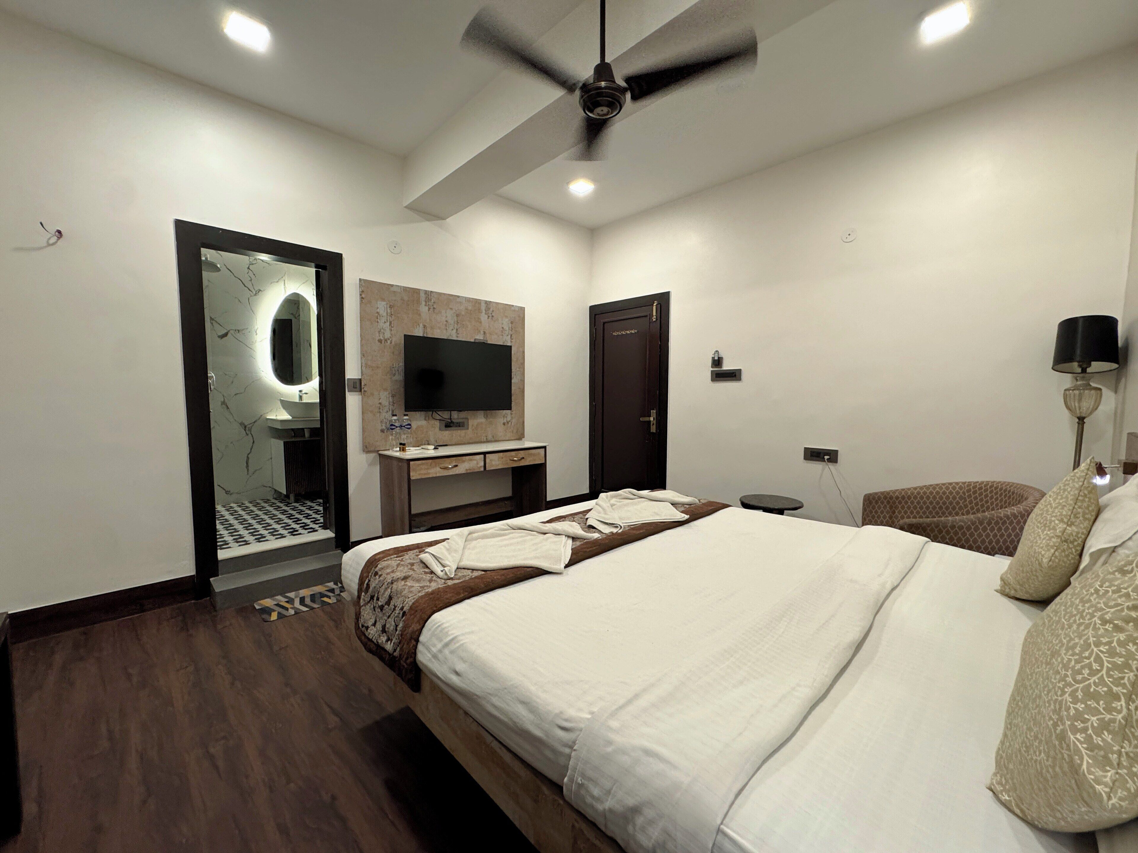 Quarto de hotel moderno com uma cama grande, uma televisão montada na parede, uma mesa com cadeira e um ventilador de teto.