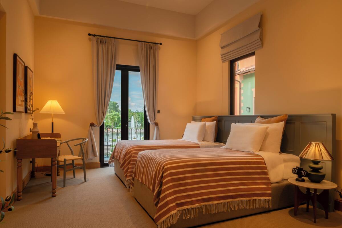 Quarto de hotel com cama, escrivaninha, cadeira, mesa pequena e janela com cortinas.