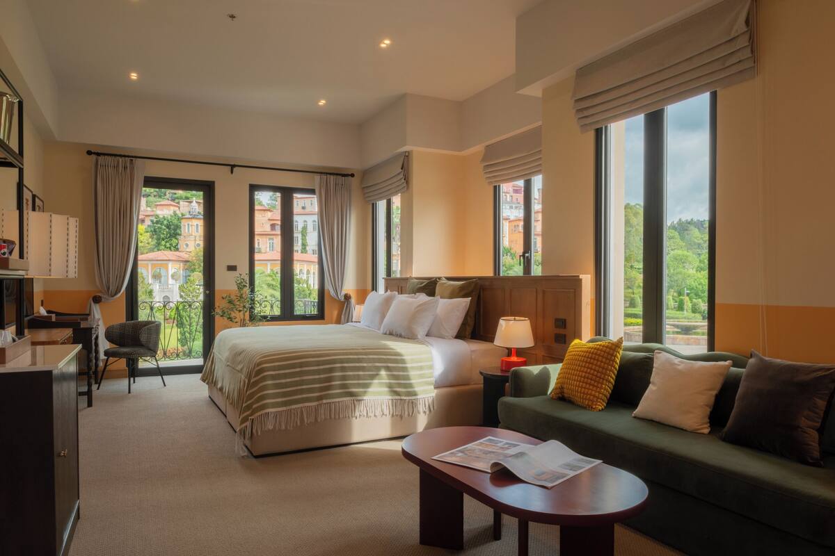 Quarto de hotel com uma cama grande, um sofá verde, uma escrivaninha e vista para edifícios pelas janelas.