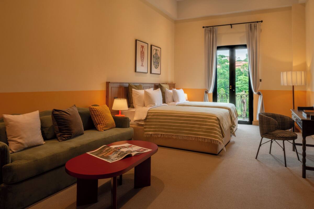 Quarto de hotel com cama, um sofá, uma mesinha com uma revista, uma escrivaninha com uma luminária e vista para o verde pela janela.