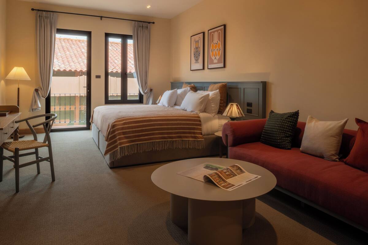 Quarto de hotel com cama, sofá vermelho, mesa de centro redonda, escrivaninha com cadeira e varanda com cortinas.