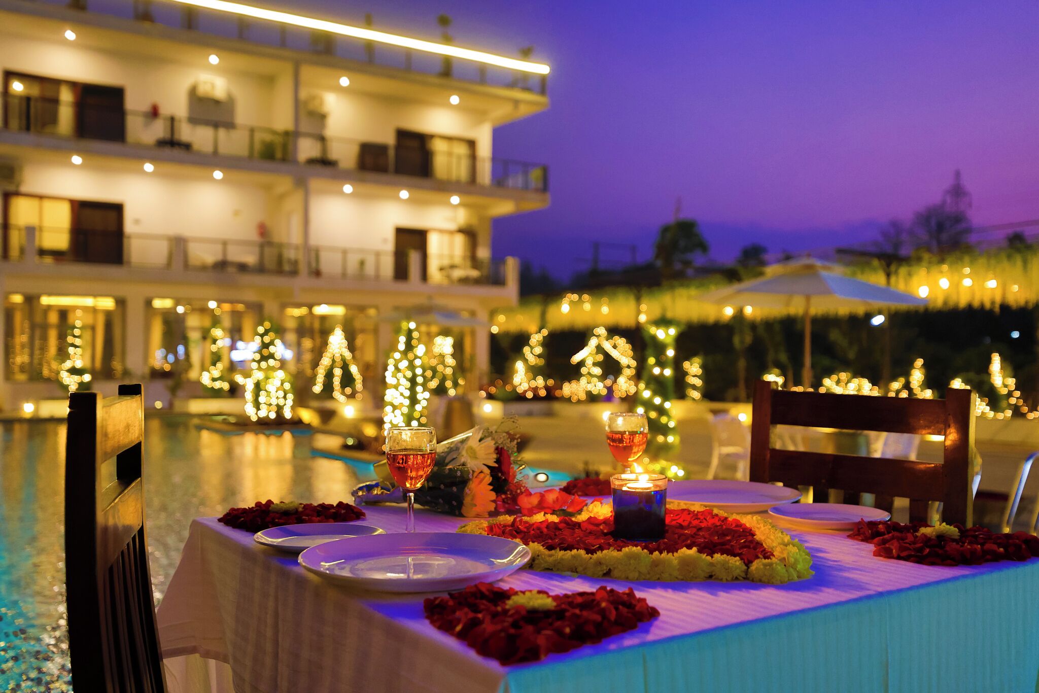 Mesa de jantar posta para dois, com pétalas de rosa, velas e copos de vinho tinto, com vista para uma piscina iluminada ao fundo.