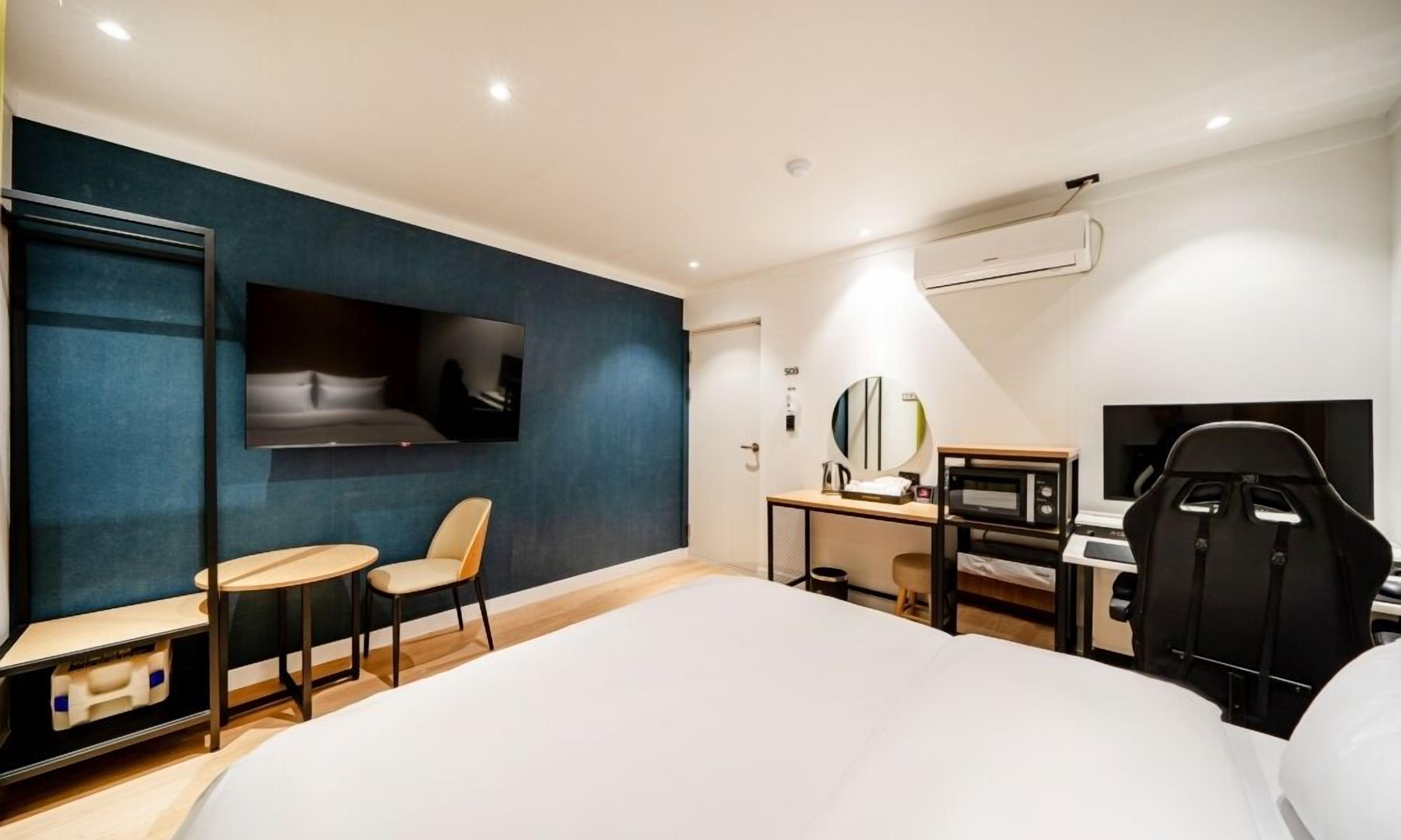 Quarto de hotel moderno com uma cama grande, uma escrivaninha com computador, uma TV na parede e uma cadeira.