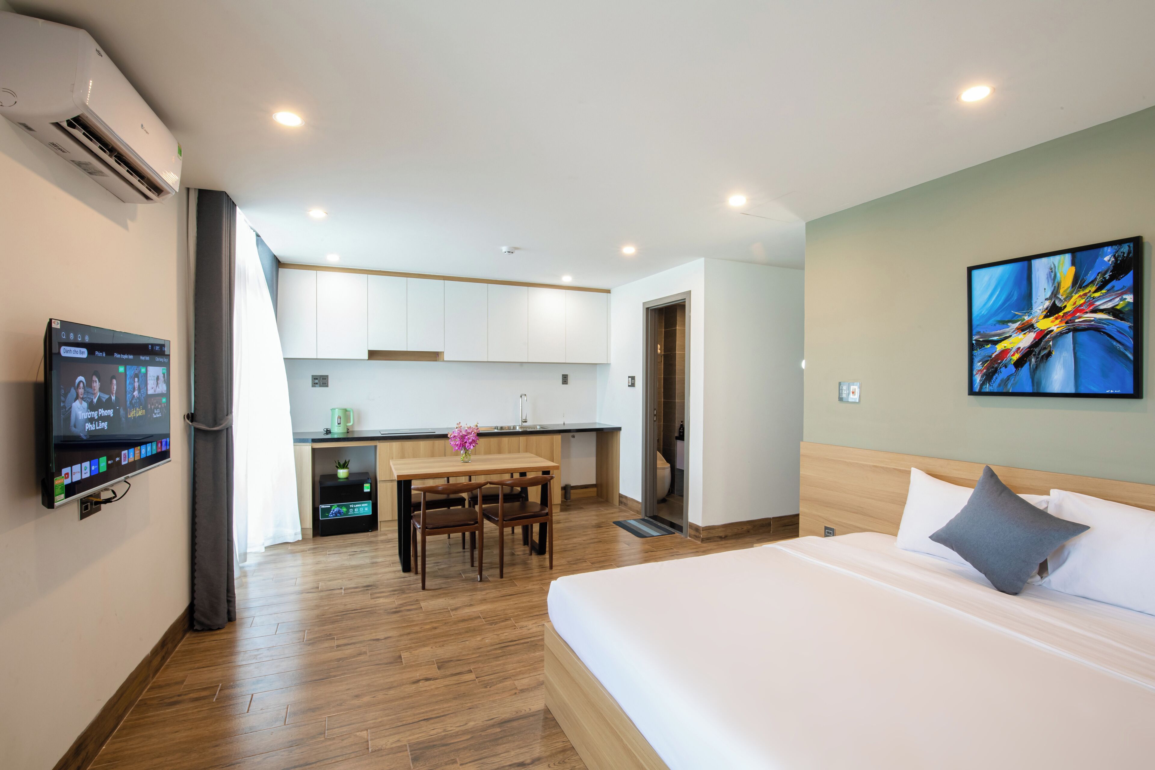 Quarto de hotel moderno com uma cama grande, área de refeições com mesa e cadeiras, cozinha e televisão montada na parede.