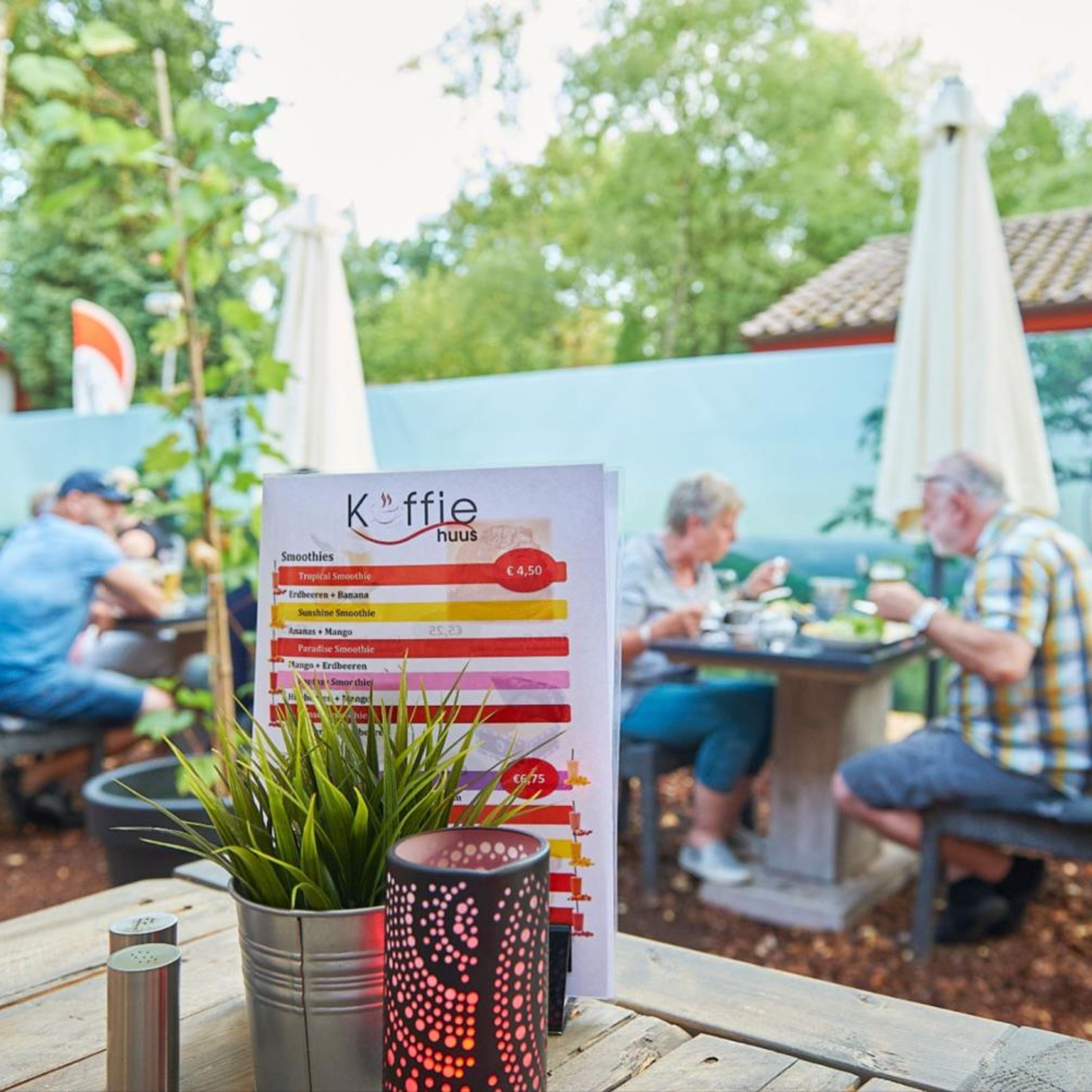 Quadro de menu para 'Koffie huis' com opções de smoothies e preços, apoiado numa mesa de madeira ao ar livre.