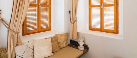 Um quarto aconchegante com teto de madeira, duas janelas com persianas de madeira, um sofá com almofadas, uma mesa de madeira e um banquinho de madeira.