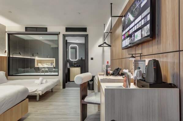 Quarto de hotel moderno com cama, escrivaninha com cafeteira, televisão e banheiro com chuveiro e pia.