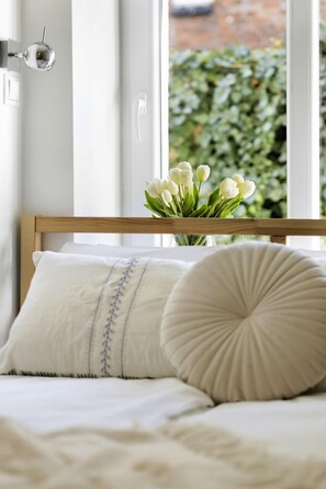 Quarto com cabeceira de madeira, cama branca com travesseiro decorativo e um vaso de flores brancas no parapeito da janela.