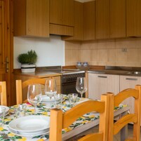 Uma cozinha com armários de madeira, uma mesa de jantar posta com pratos e copos, e um vaso de planta no balcão.