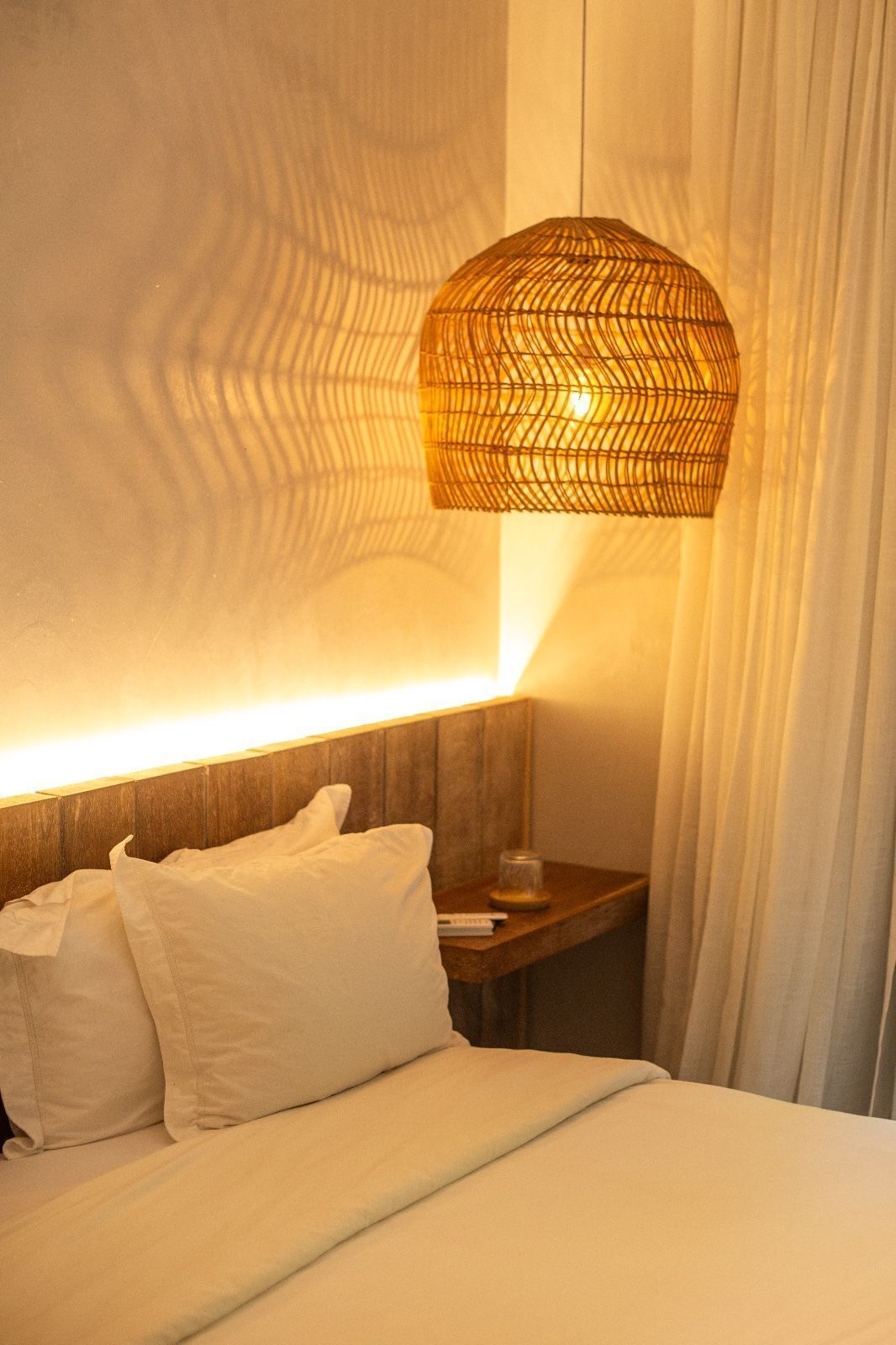 Uma cama bem arrumada com lençóis brancos, cabeceira de madeira e uma luminária trançada em cima.