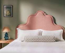 Uma cama bem arrumada com cabeceira rosa texturizada, almofadas brancas e uma almofada estampada. Uma luminária azul e um copo de água estão em uma mesa de cabeceira de madeira ao lado da cama.