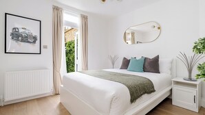 Quarto com cama branca, radiador, quadro emoldurado, espelho e mesa de cabeceira com planta.