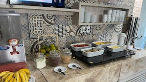 Disposição de buffet com diversos alimentos, uma exposição de frutas e um dispensador de bebidas.