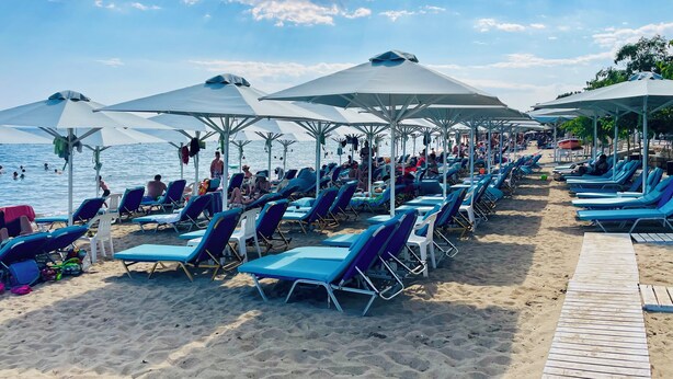 Uma praia com fileiras de espreguiçadeiras e guarda-sóis, um calçadão de madeira e pessoas nadando na água.