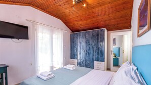 Quarto com teto de madeira, cama com roupa de cama branca e azul, televisão montada na parede e guarda-roupa com porta azul.
