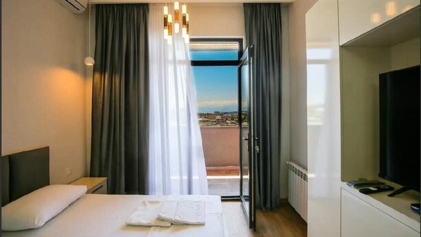 Quarto de hotel com cama, escrivaninha, televisão e varanda com cortinas.