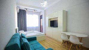 Quarto de hotel moderno com sofá, cama, mesa de jantar com cadeiras, televisão e cozinha compacta.