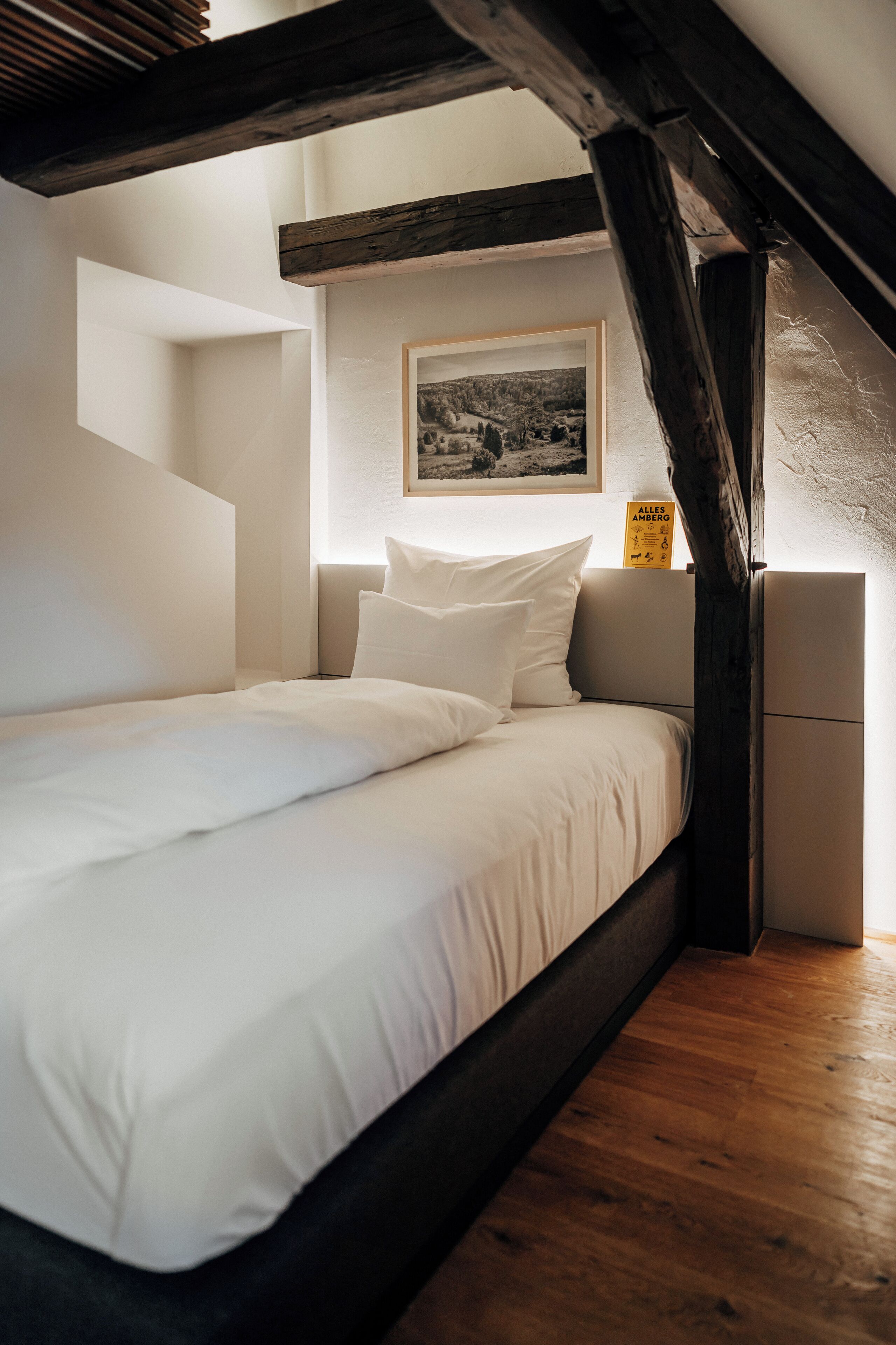 Quarto com teto de madeira, cama com roupa de cama branca, mesa de cabeceira e um quadro na parede.