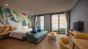 Quarto de hotel com cama, sofá, televisão, mesa de jantar e vista para o mar.