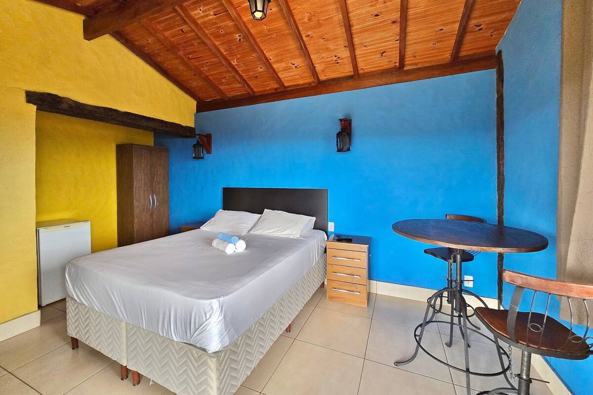 Quarto com cama, mesa de cabeceira e uma pequena mesa redonda com uma cadeira.