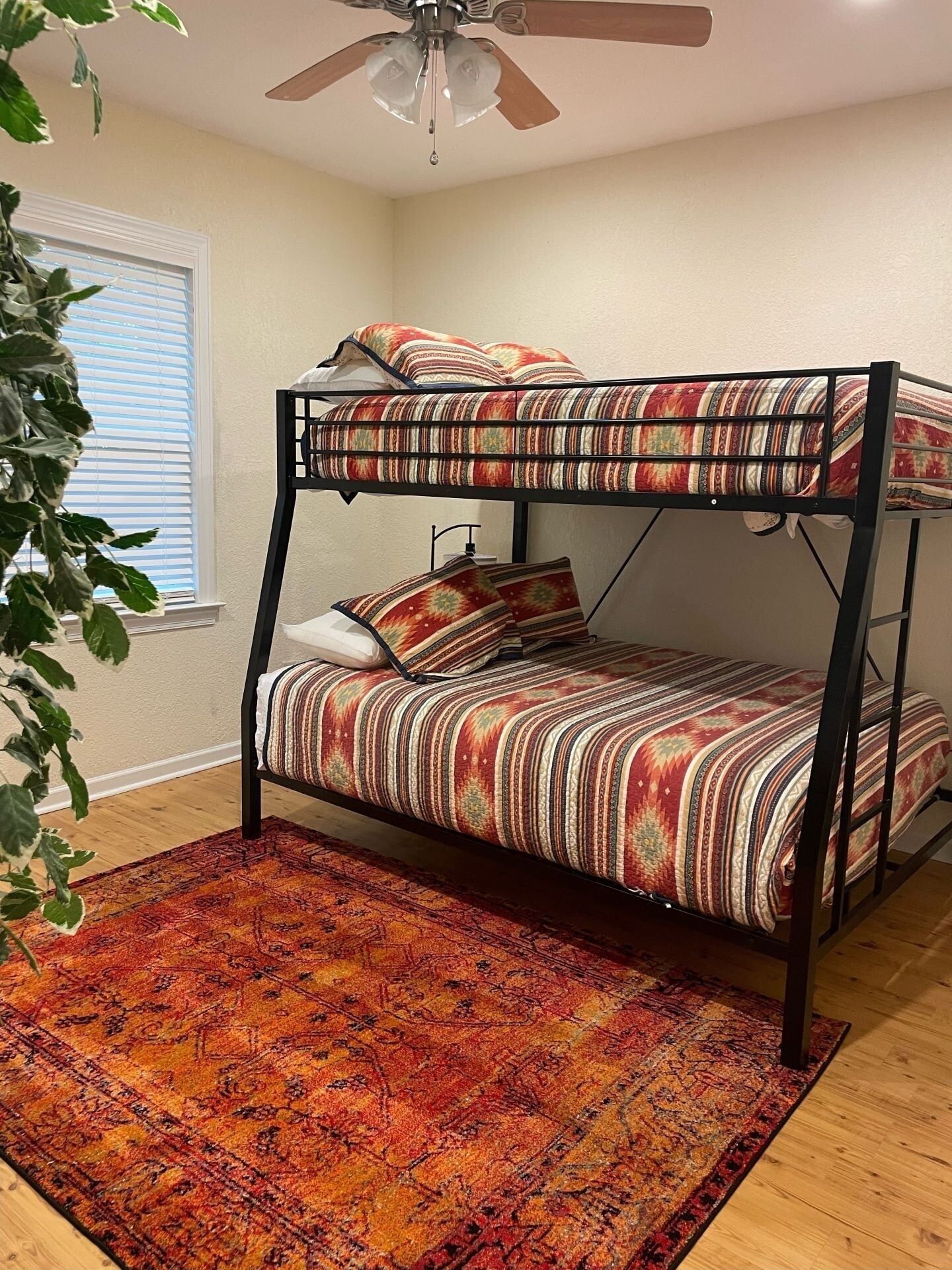 Uma cama beliche com roupa de cama estampada, um ventilador de teto, uma janela com persianas e um tapete decorativo.
