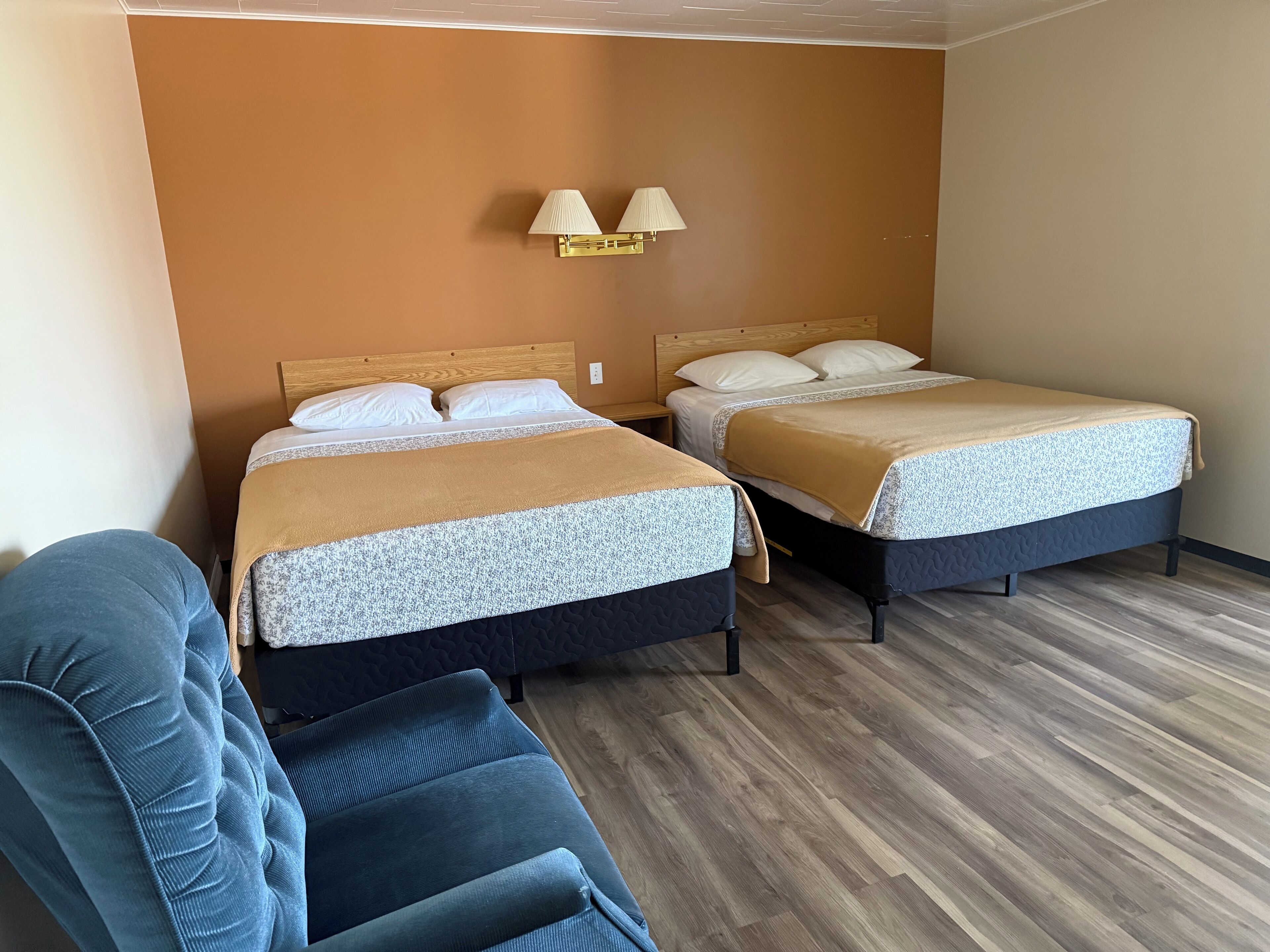 Quarto de hotel com duas camas, uma mesa de cabeceira, uma luminária e uma cadeira azul.