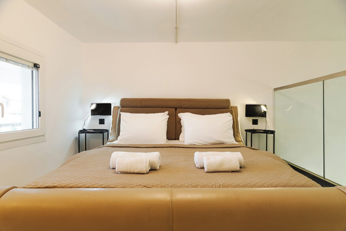 Uma cama bem arrumada com cabeceira marrom, duas mesas de cabeceira com abajures e uma janela com persianas.