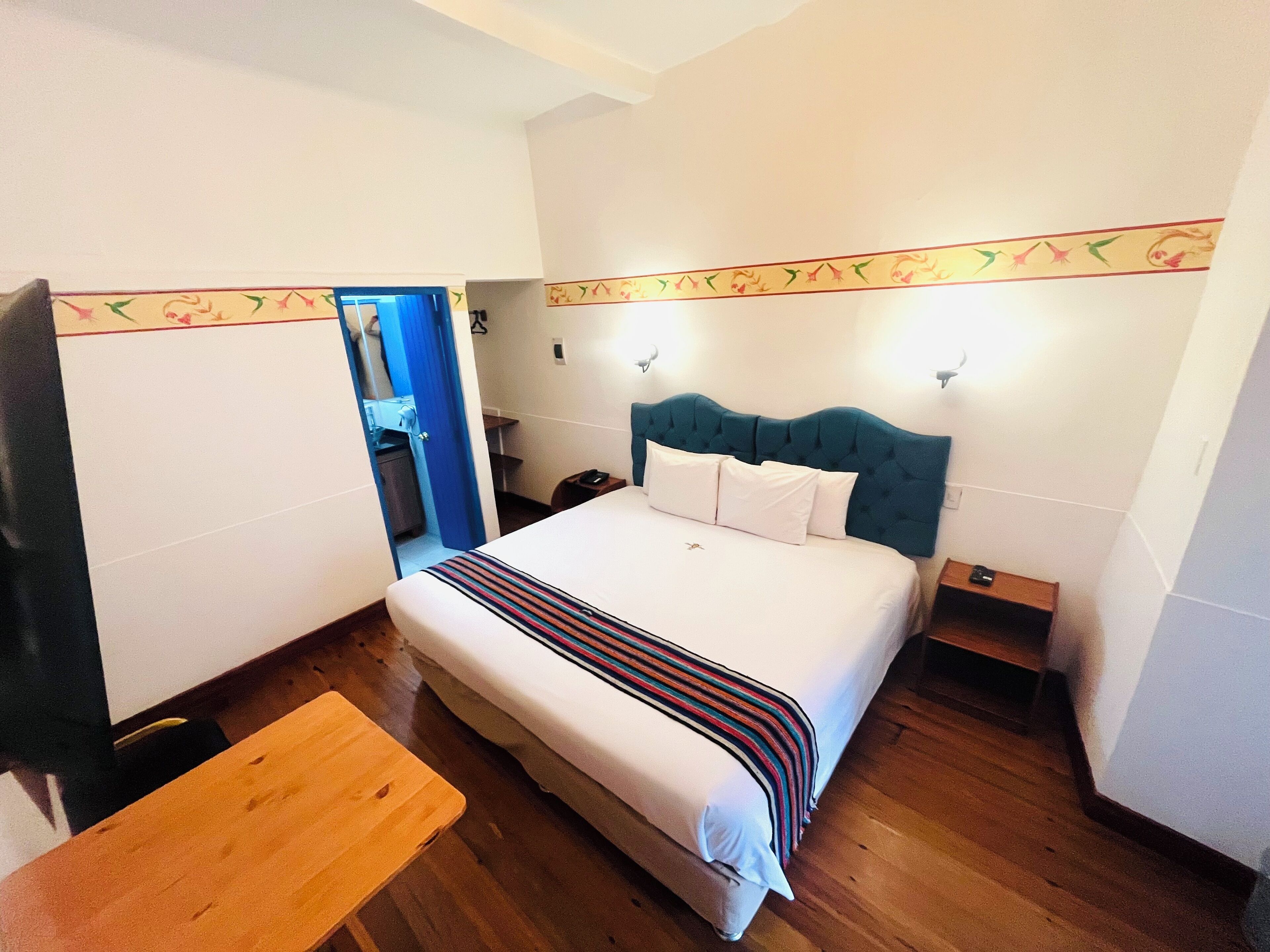 Quarto de hotel com uma cama, mesas de cabeceira, um banco de madeira e um banheiro visível por uma porta aberta.
