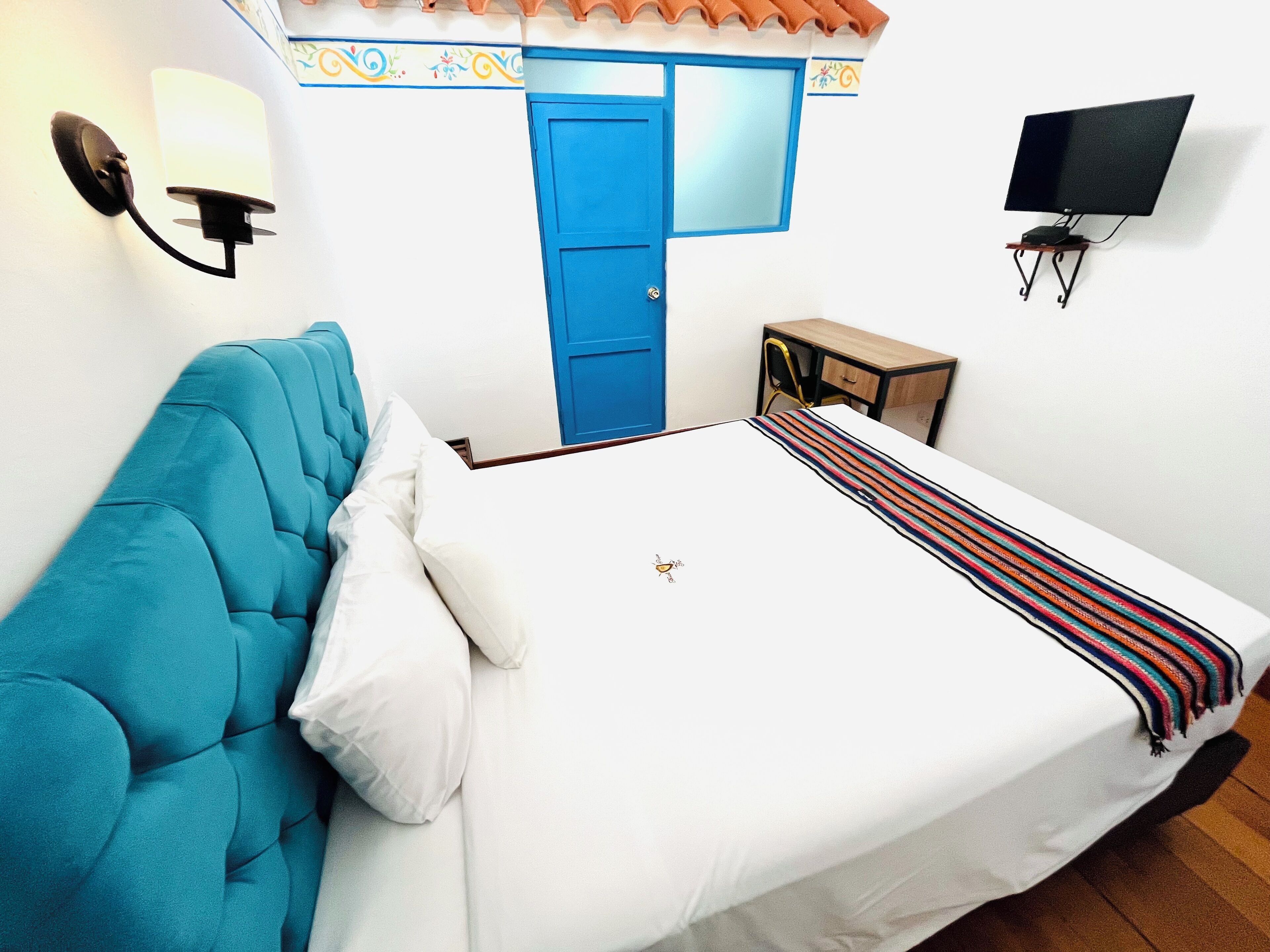 Quarto com cabeceira estofada azul, colcha branca, manta listrada colorida, mesa de cabeceira de madeira pequena, escrivaninha com cadeira, porta azul, televisão montada na parede e luminária de parede.