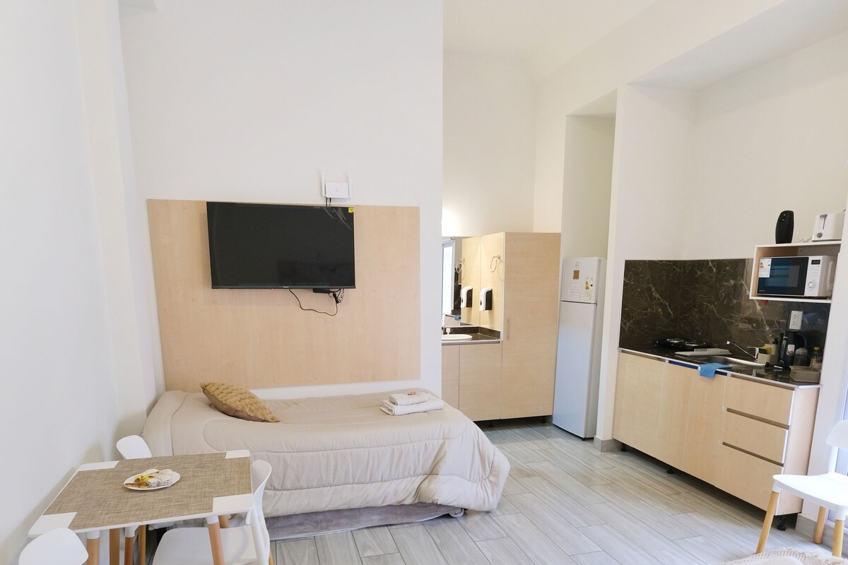 Quarto de hotel moderno com mesa de jantar, duas camas, TV e cozinha, além de TV de tela plana na parede.