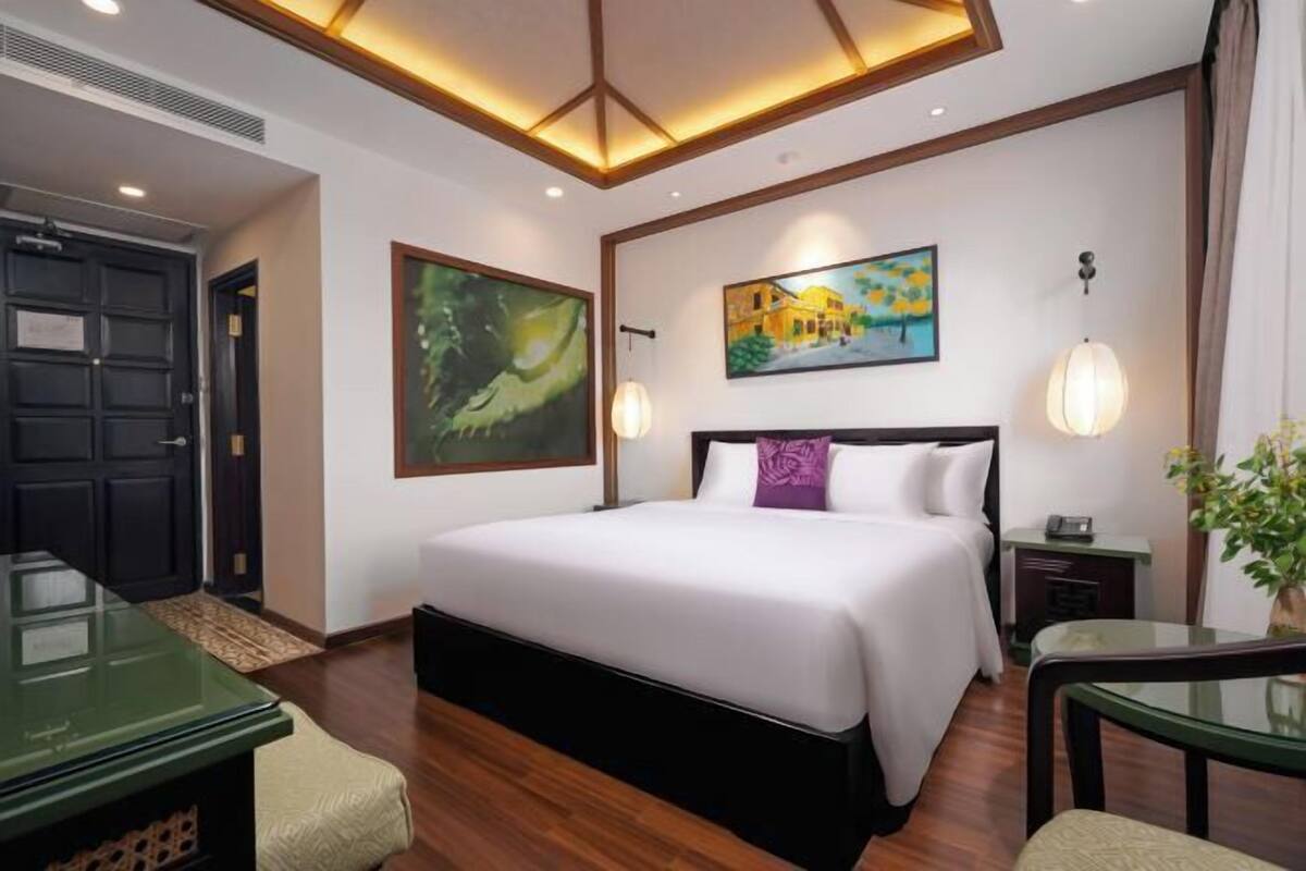 Quarto de hotel com cama, escrivaninha, cadeira, mesa de cabeceira, luminária e um quadro na parede.