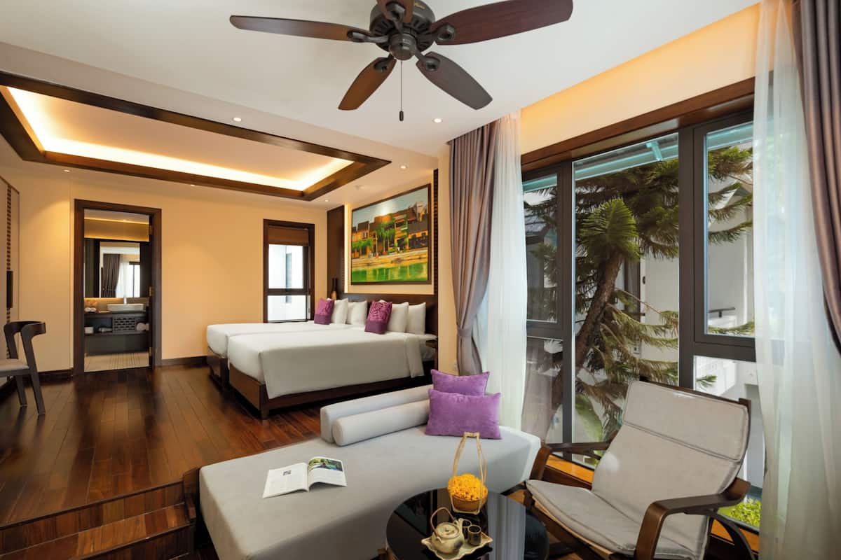 Quarto de hotel com cama, sofá, poltrona, ventilador de teto, janela com vista para palmeiras e um quadro na parede.