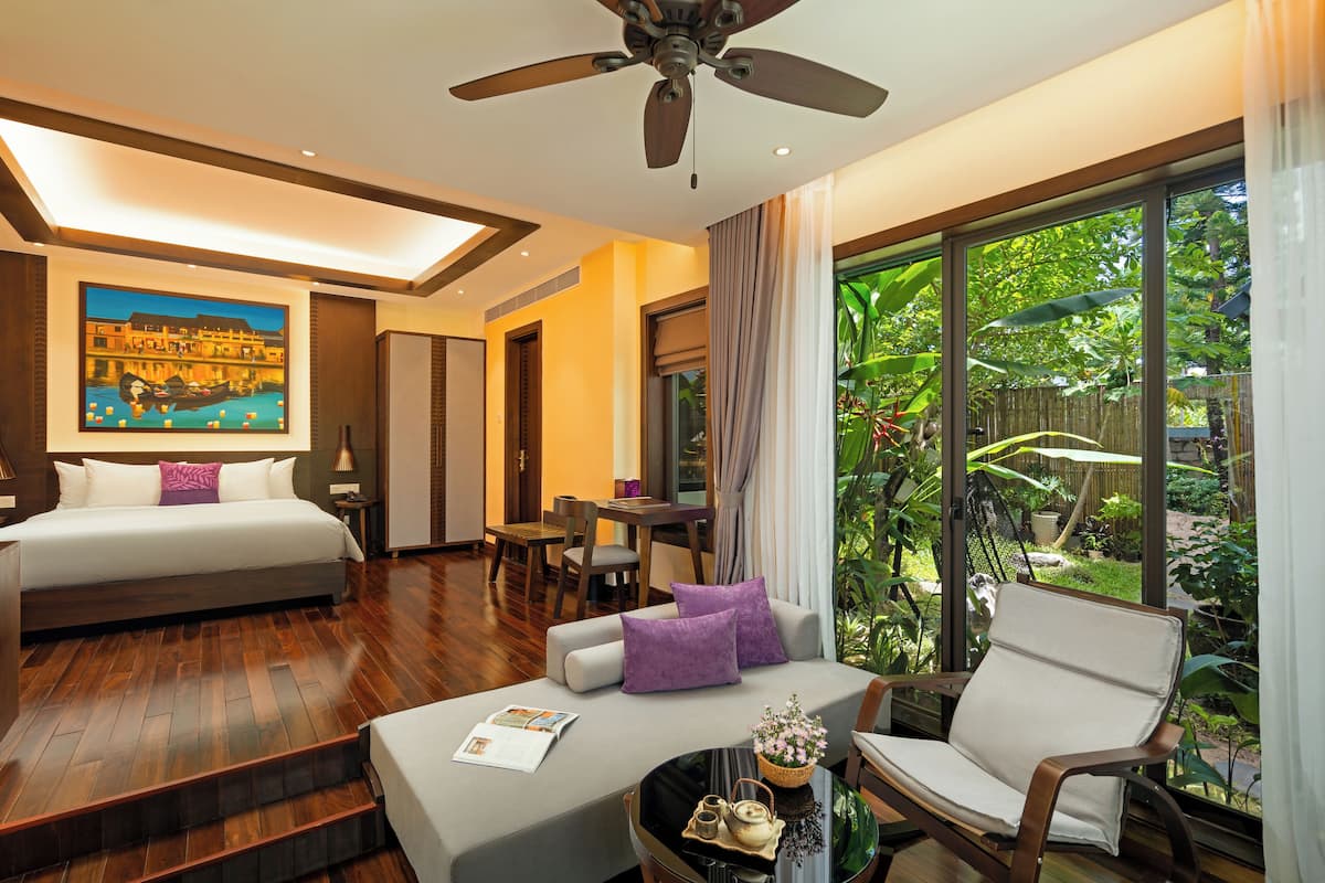 Quarto de hotel com cama, sofá, mesa de jantar e uma cadeira. Há uma janela grande com vista para o verde lá fora.
