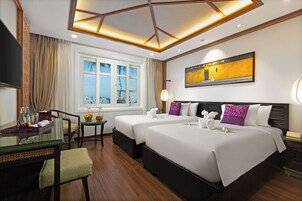Quarto de hotel com duas camas, uma escrivaninha com cadeira, janela com cortinas, um quadro na parede e luminária no teto.