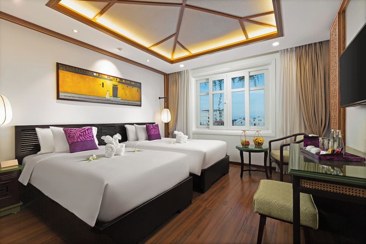 Quarto de hotel com duas camas, uma escrivaninha, uma cadeira, janela com vista e um quadro na parede.