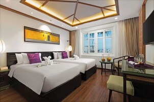 Quarto de hotel com duas camas, uma escrivaninha, uma cadeira, janela com vista e um quadro na parede.