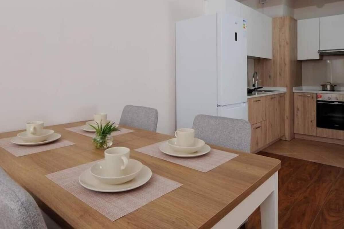 Uma área de refeições com mesa de madeira posta para quatro pessoas, geladeira e cozinha com armários de madeira e fogão.