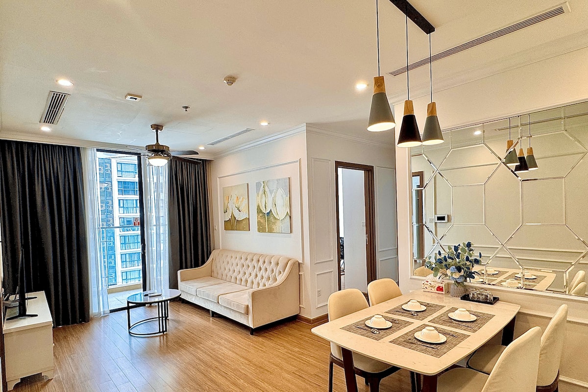 Uma sala de estar moderna com sofá, mesa de jantar e cadeiras. Há uma janela com cortinas, um ventilador de teto e obras de arte na parede.