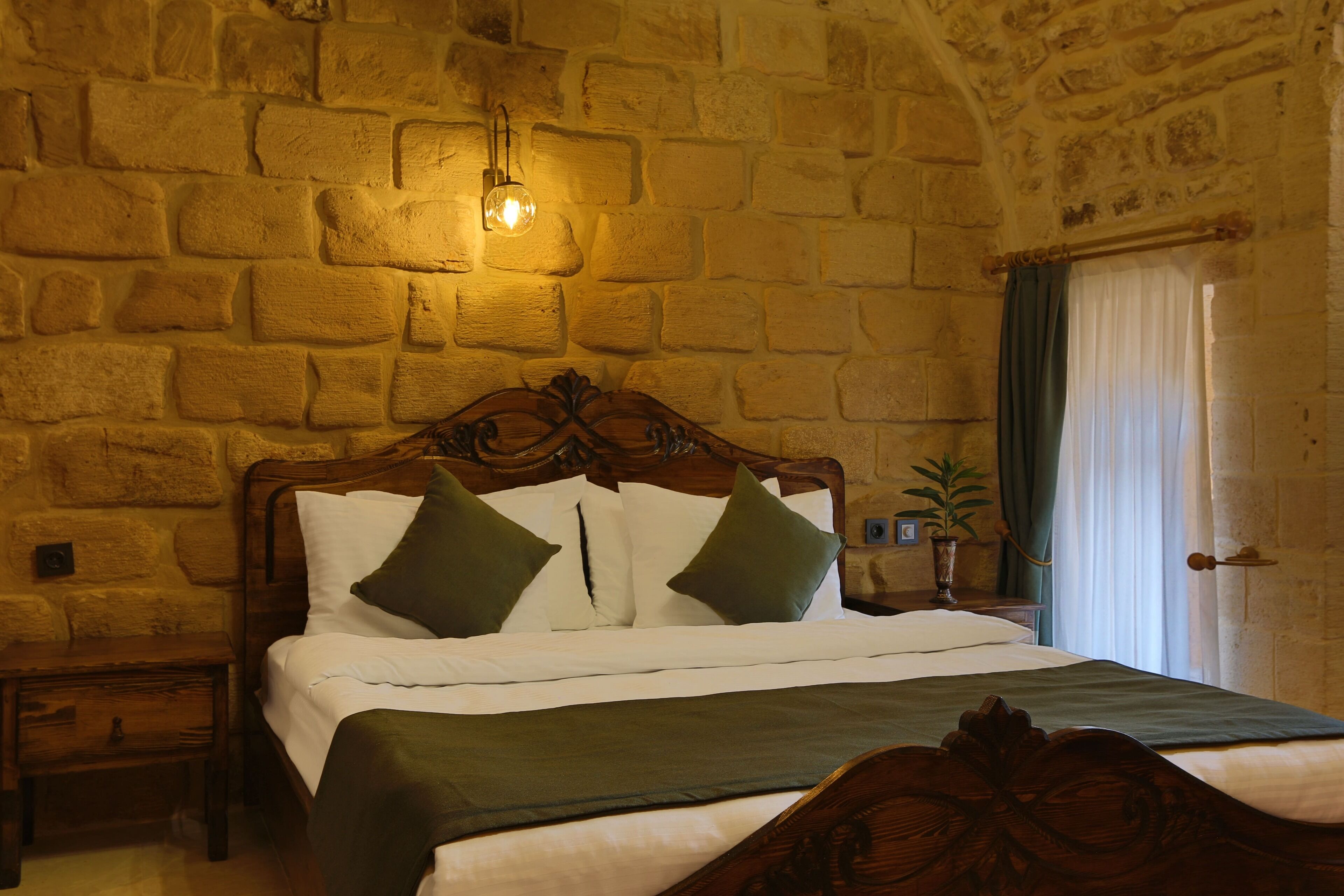 Quarto com parede de pedra, cama de madeira com roupa de cama verde e branca, mesa de cabeceira e planta em vaso.