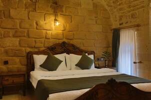 Quarto com parede de pedra, cama de madeira com roupa de cama verde e branca, mesa de cabeceira e planta em vaso.