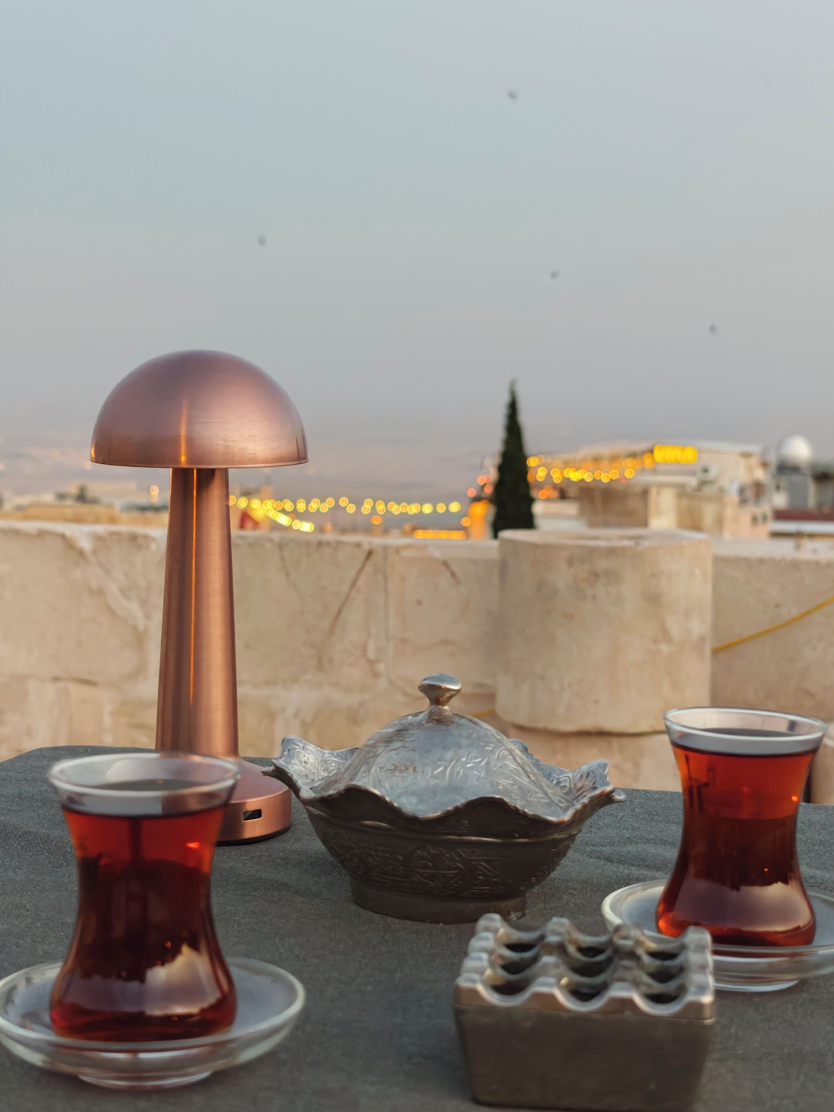 Uma mesa com uma luminária de cobre, um suporte de metal e duas taças de vidro com líquido vermelho, ao fundo de uma vista urbana.