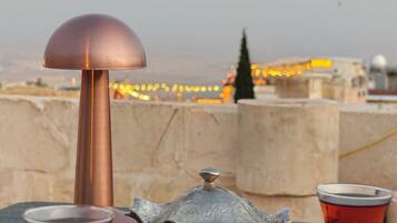 Uma mesa com uma luminária de cobre, um suporte de metal e duas taças de vidro com líquido vermelho, ao fundo de uma vista urbana.