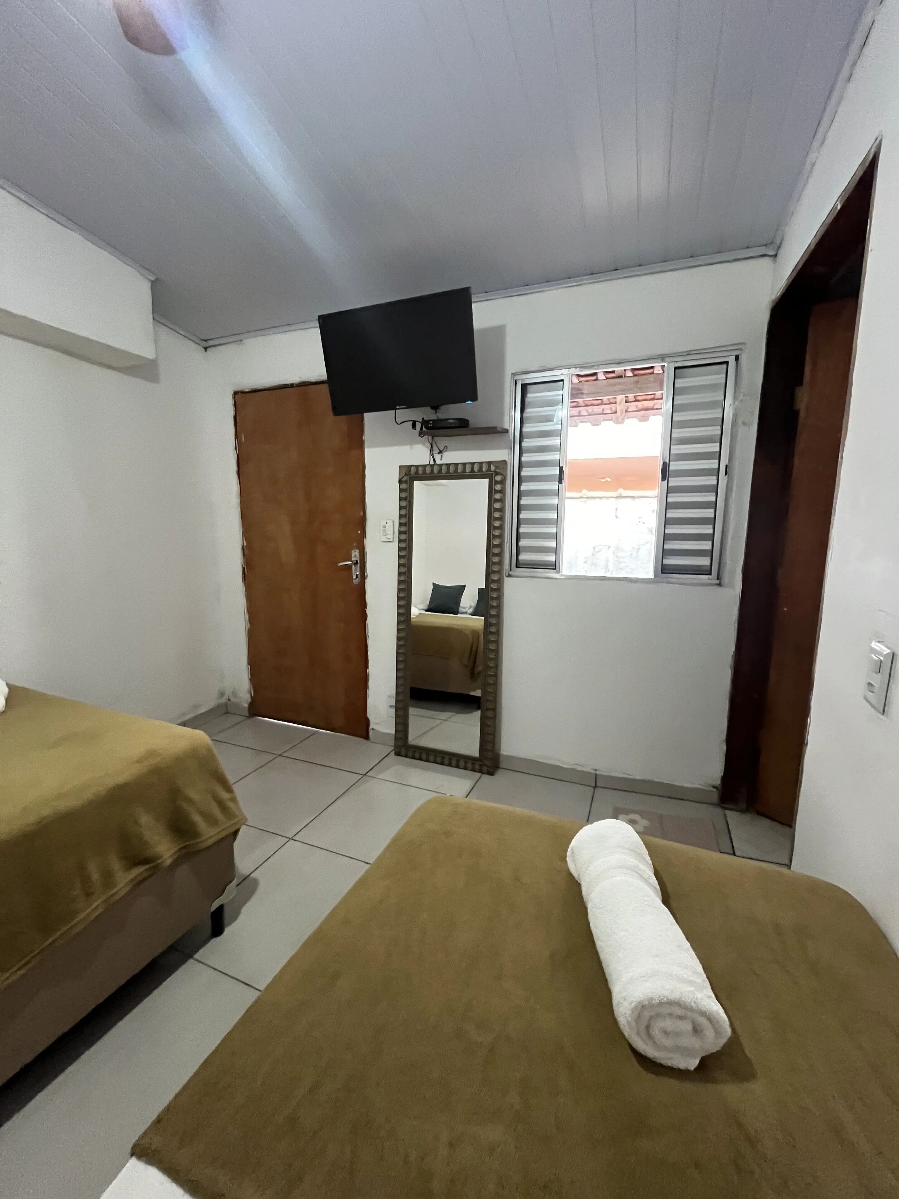 Quarto com duas camas, televisão na parede, espelho e janela com persianas.