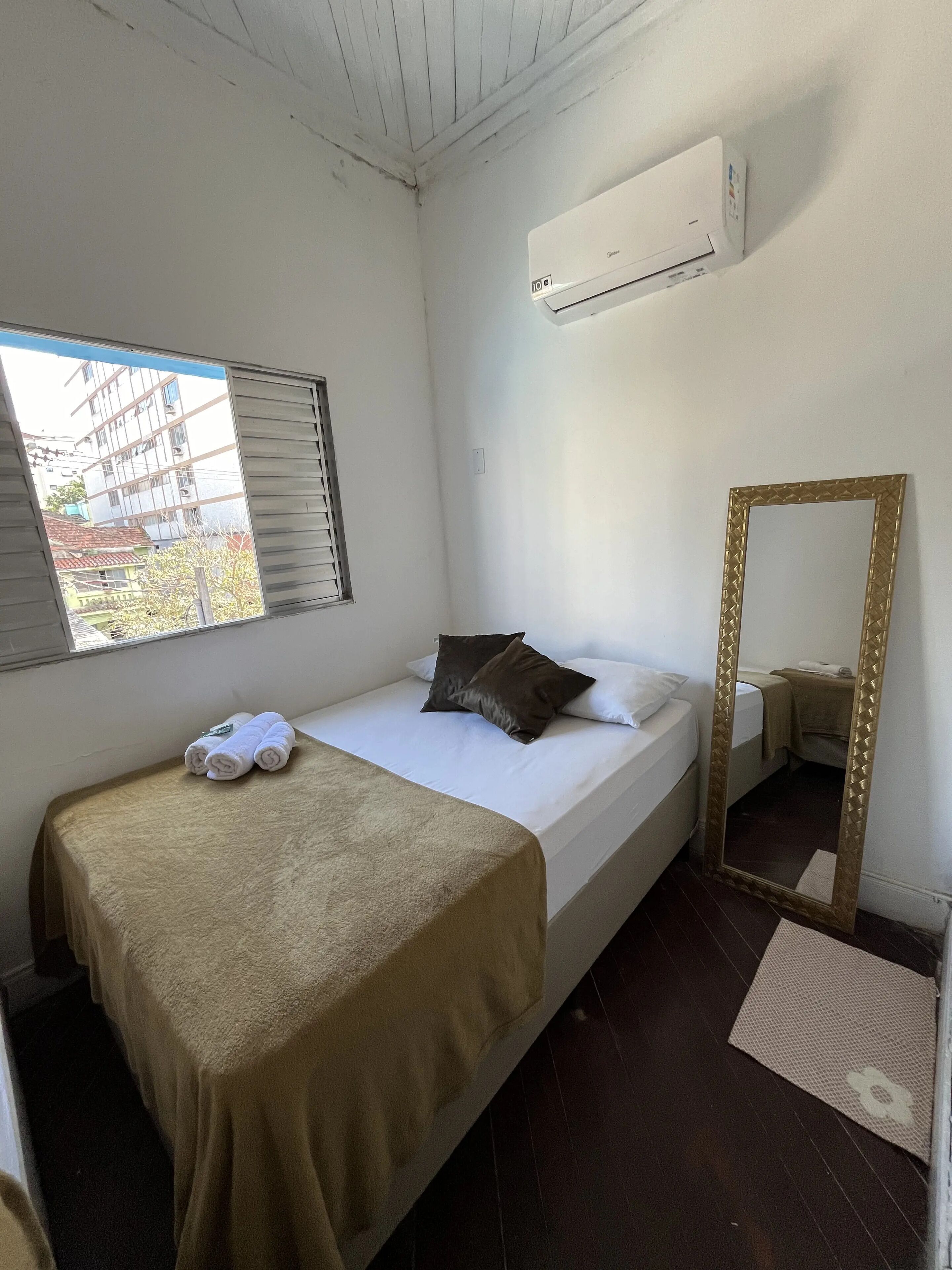Quarto pequeno e limpo com uma cama de solteiro, janela com persianas, ar condicionado e um espelho.